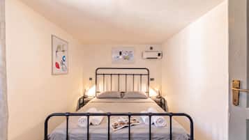 Uma cama de solteiro com estrutura de metal, duas luminárias de cabeceira, um ar condicionado fixado na parede e duas obras de arte emolduradas.