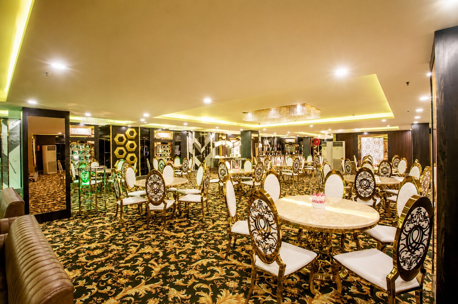 Espaço de refeições de hotel com mesas redondas e cadeiras elaboradas, piso coberto por carpete e um candelabro.