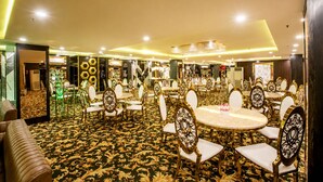 Espaço de refeições de hotel com mesas redondas e cadeiras elaboradas, piso coberto por carpete e um candelabro.