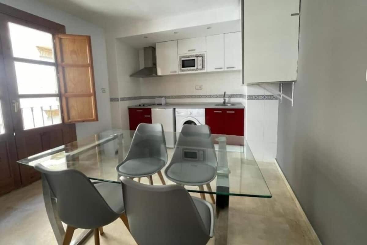 Uma cozinha moderna com mesa de jantar de vidro, cadeiras cinzas, armários brancos e bancada vermelha.