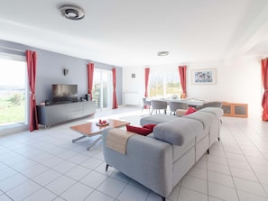 Uma sala de estar moderna com um sofá cinzento, uma TV, uma área de jantar com cadeiras e grandes janelas com cortinas.