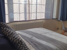 Um quarto com cama, radiador, janela com cortinas e uma cadeira.