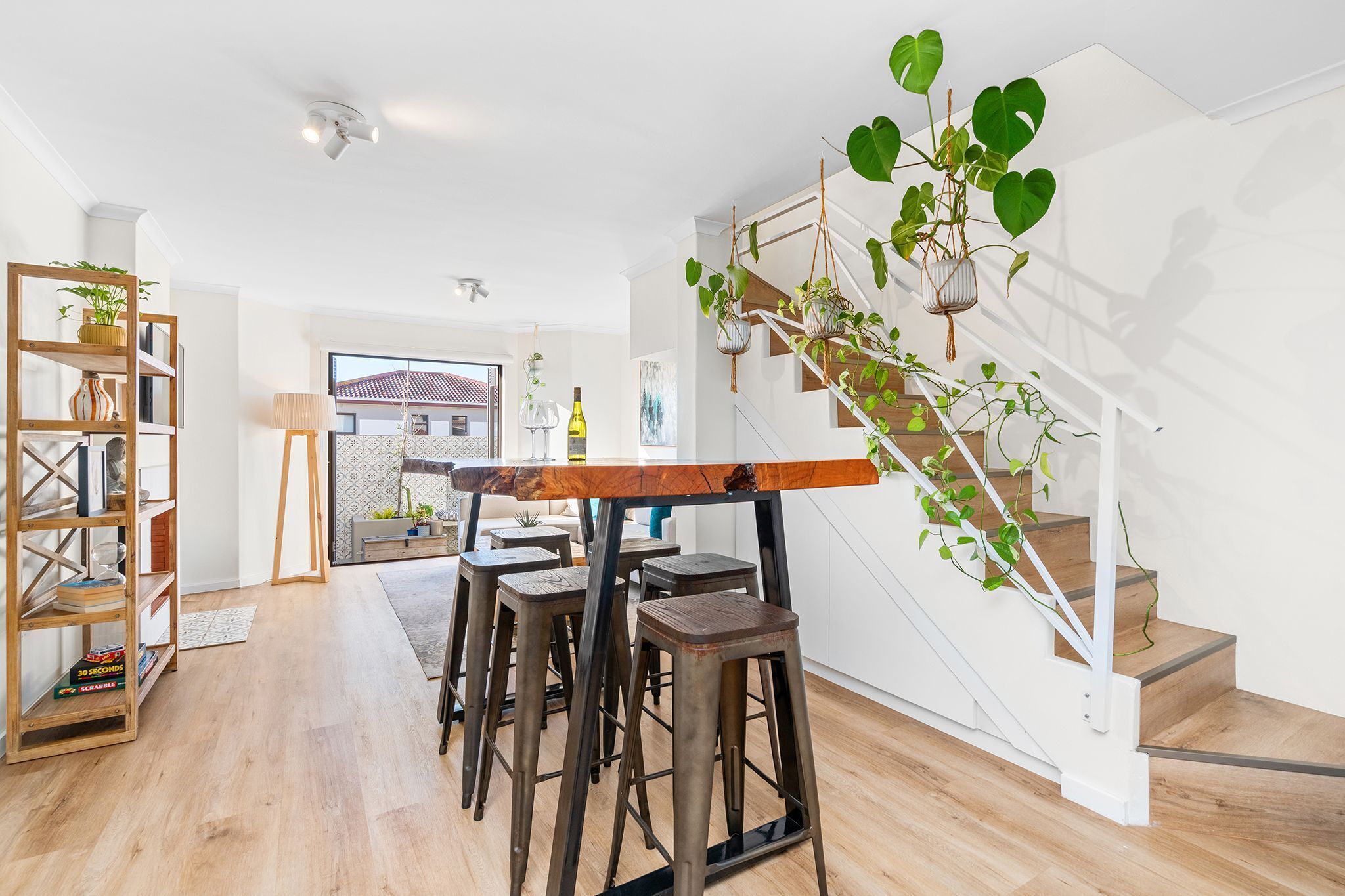 Um interior moderno com uma mesa de jantar de madeira, banquetas de bar, uma escada e plantas penduradas.