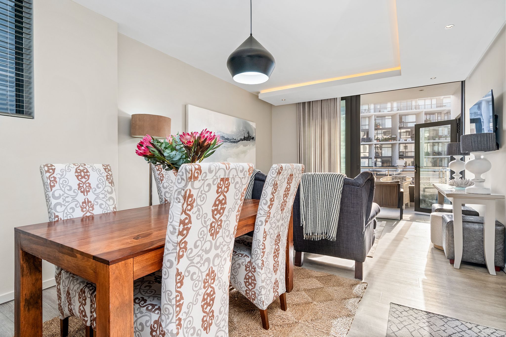Uma sala de jantar moderna com uma mesa de madeira, cadeiras estampadas, um vaso de flores e uma luminária pendente.