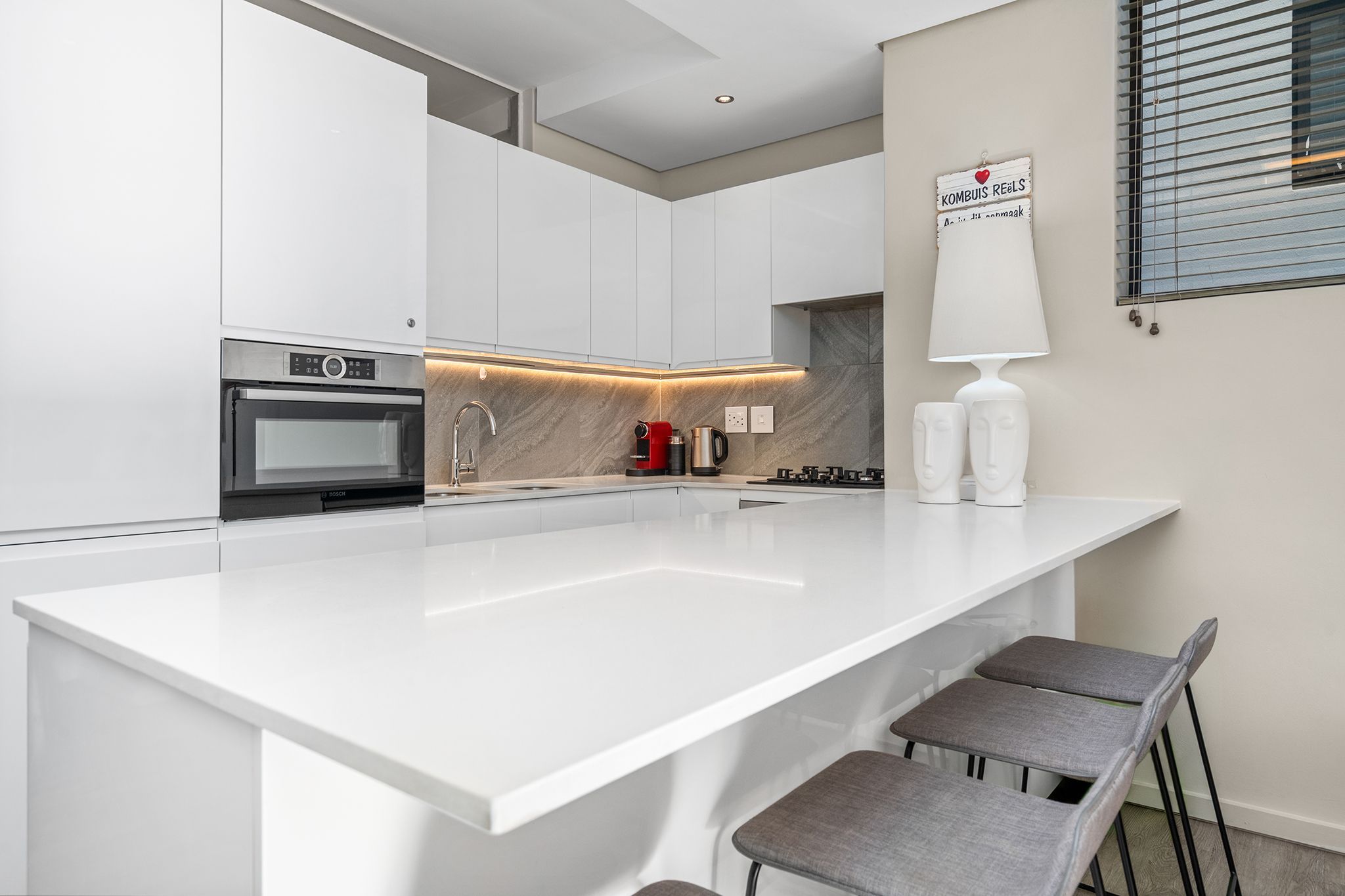Cozinha moderna com armários brancos, forno Bosch, cafeteira e painel de fundo em mármore.