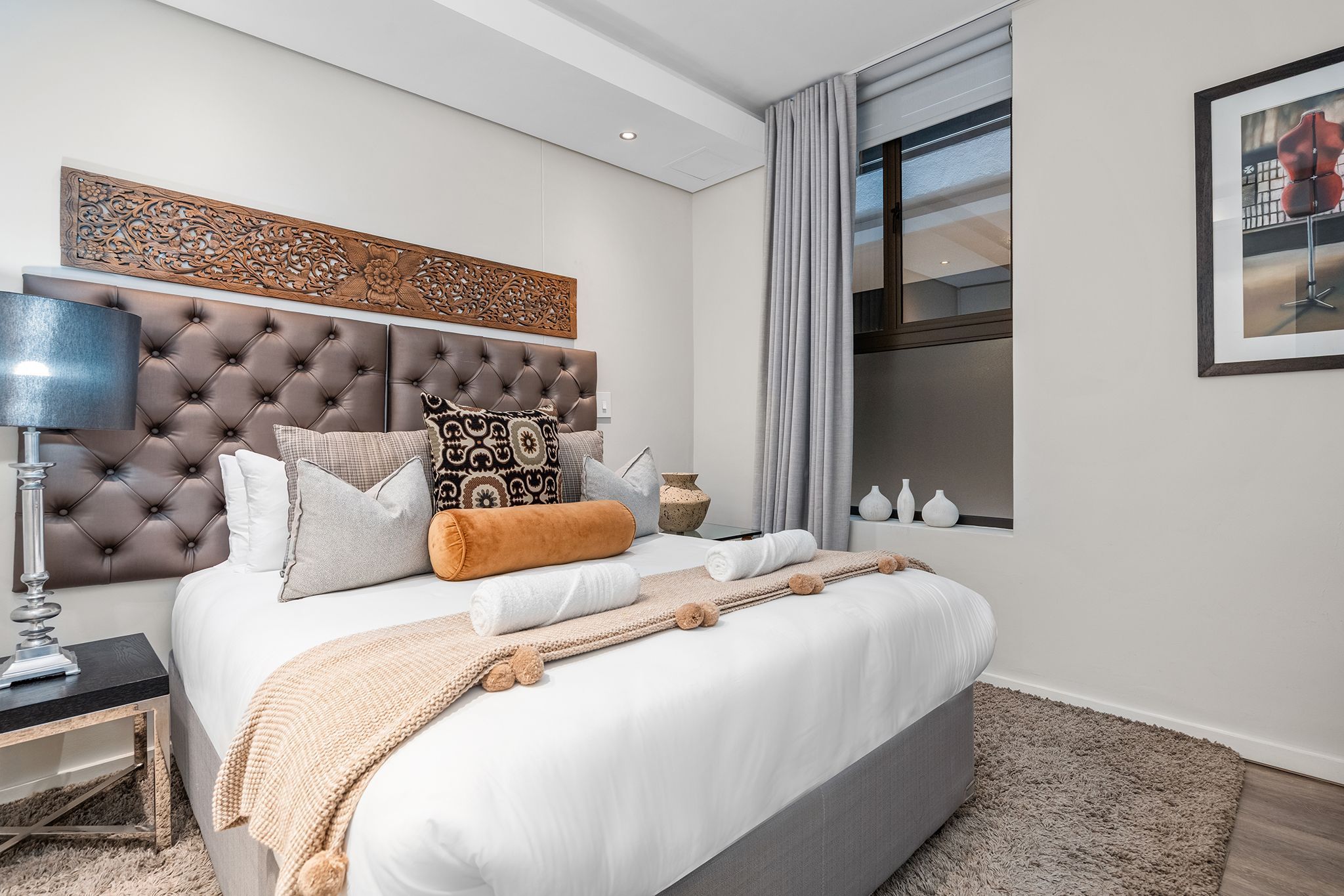 Quarto bem arrumado com cabeceira estofada, mesa de cabeceira com abajur, janela grande com cortinas e uma obra de arte emoldurada na parede.