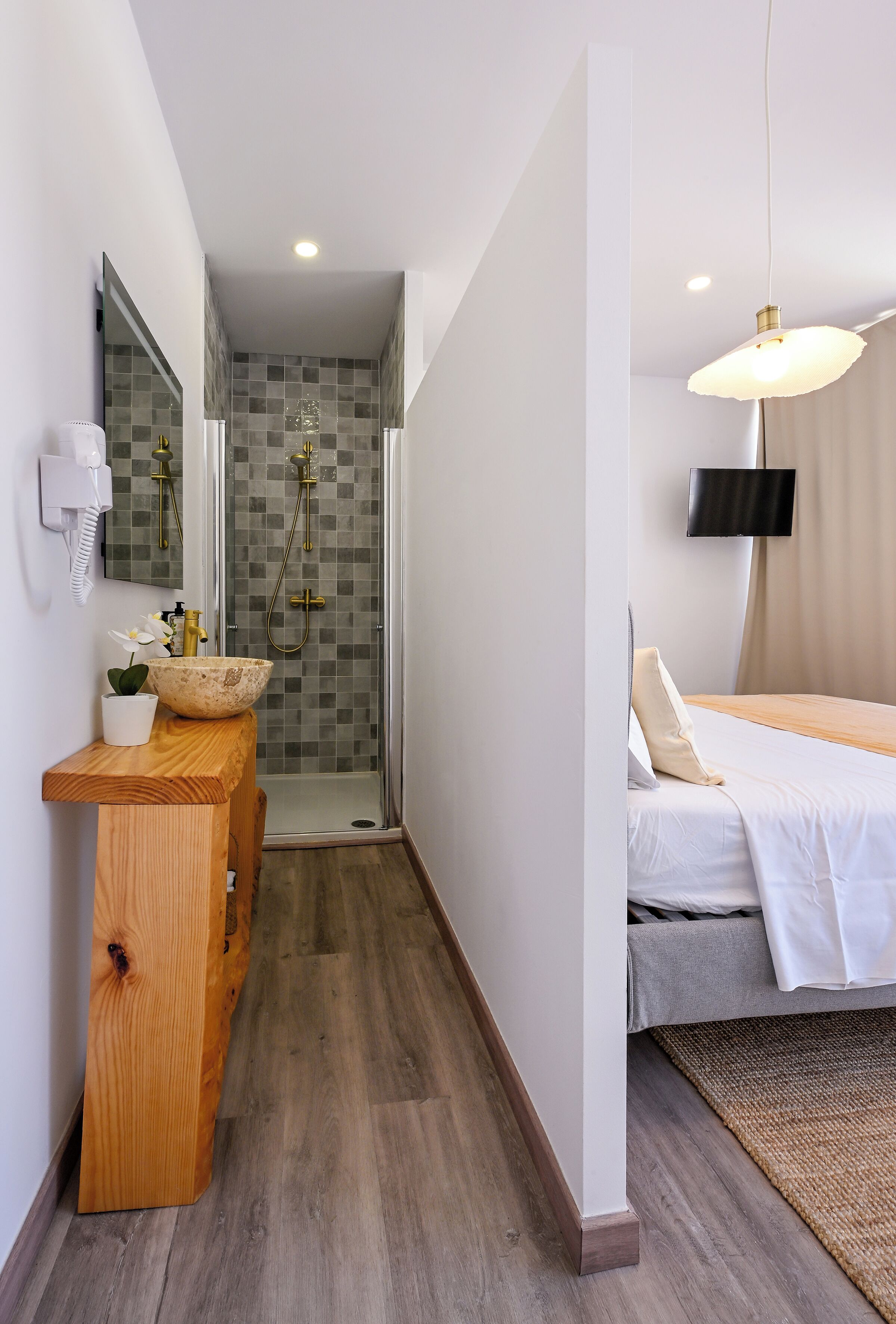 Quarto de hotel com toucador de madeira, chuveiro com porta de vidro, cama com roupa de cama branca e televisão montada na parede.