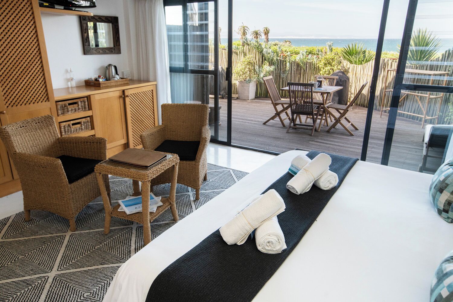 Quarto de hotel com uma cama, duas cadeiras de vime, uma mesinha, um armário de madeira e vista para o oceano.