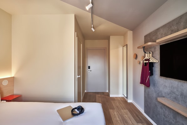 Quarto de hotel com cama, escrivaninha, televisão na parede e corredor que dá para outro quarto.