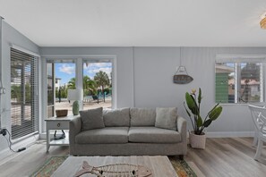 Uma sala de estar com um sofá, uma mesinha, uma luminária, uma planta em vaso e vista para o exterior.