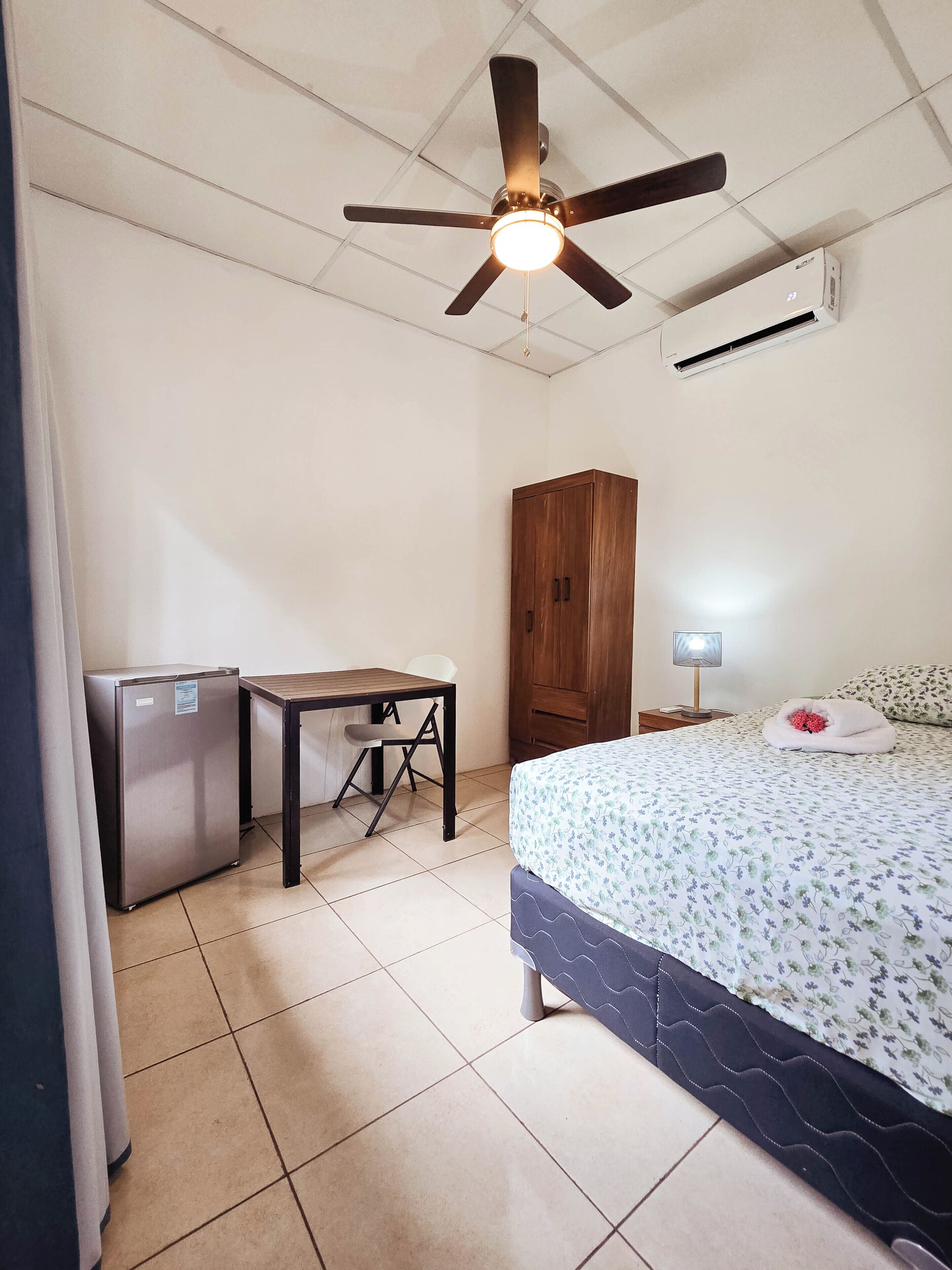 Quarto com cama, escrivaninha, guarda-roupa, ventoinha no teto e ar condicionado.