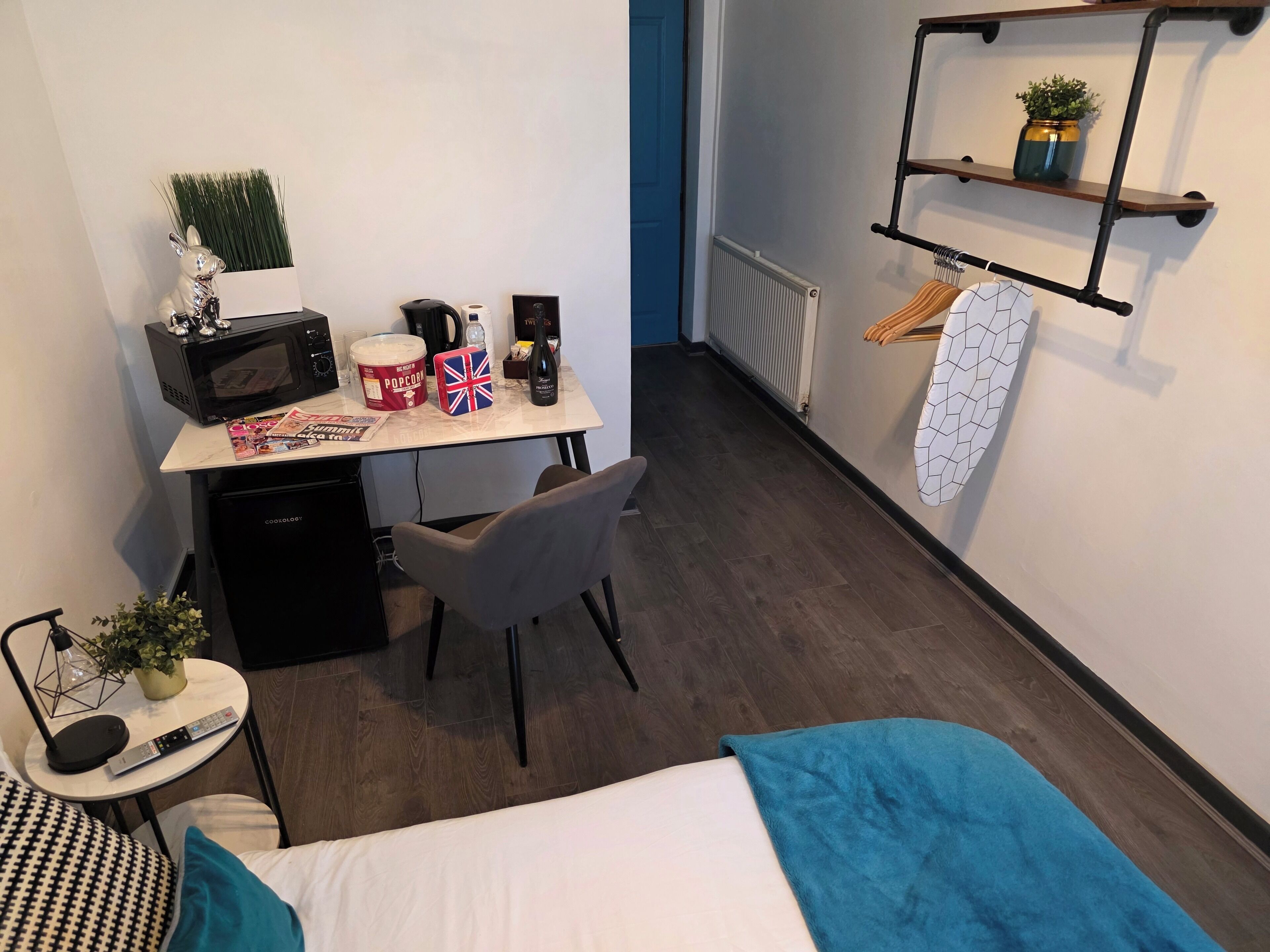 Quarto pequeno com cozinha compacta, uma cama, uma cadeira, uma mesinha com um controle remoto e uma prateleira com uma planta.