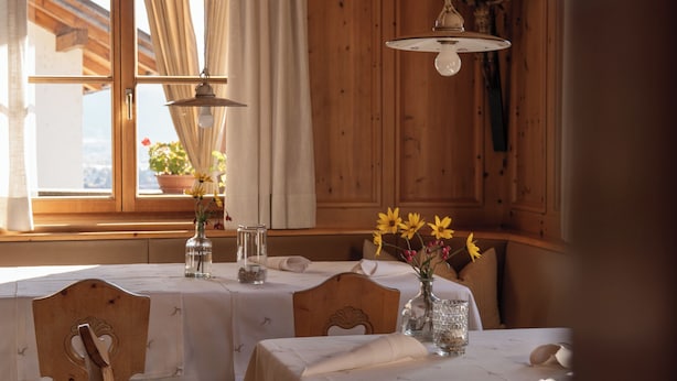 Interior de madeira com mesa de jantar posta para dois, cadeiras, luminárias pendentes e uma janela com cortinas.