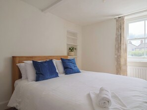 Cama bem arrumada com roupa de cama branca e azul, cabeceira de madeira, uma pequena planta em vaso em uma prateleira e uma janela com cortinas.