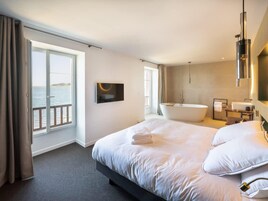 Quarto de hotel moderno com cama grande, banheira, janela ampla com vista para o mar e decoração minimalista.