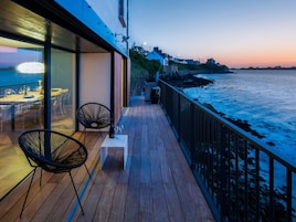 Uma varanda com vista para o mar, área de refeições com cadeiras e uma mesa com garrafa e copos.