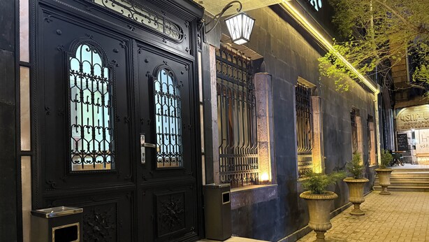 Uma entrada negra e ornamentada com portões de metal, um banco de pedra e um caminho pavimentado que leva a um edifício.