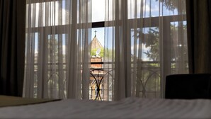 Quarto com cama, janela com cortinas e vista para um edifício com cruz no topo.