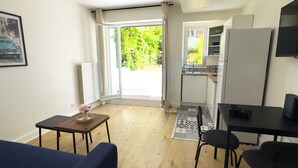 Uma sala moderna com sofá, uma mesinha com um vaso, área de refeições com cadeiras, cozinha com geladeira e uma porta de vidro deslizante para uma varanda.