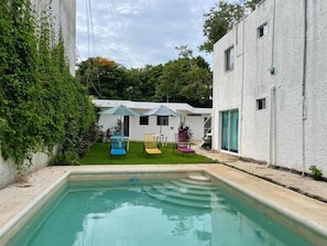 Uma piscina com uma pequena área de deck, um edifício branco com telhado plano e um gramado verde com mobiliário de exterior.