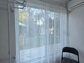 Quarto com uma grande janela coberta por cortinas finas, uma cadeira preta e um aparelho de ar condicionado na parede.