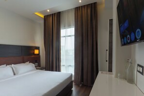 Quarto de hotel com cama, mesa de cabeceira, televisão e janela com cortinas.