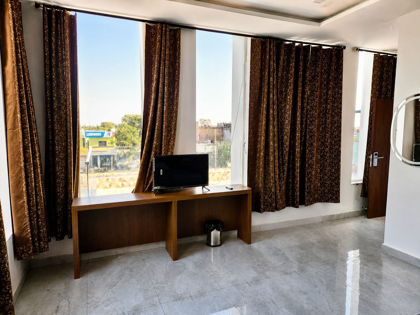 Quarto com televisão sobre suporte de madeira, janelas amplas com cortinas, piso ladrilhado e porta com espelho redondo.