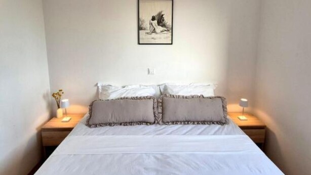 Quarto com cama, duas mesas de cabeceira com luminárias, um quadro na parede e um pequeno vaso de flores em uma das mesas.