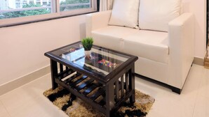 Sala de estar com sofá branco, mesa de centro de vidro com uma planta, relógio de parede redondo e vista para edifícios através de uma grande janela.
