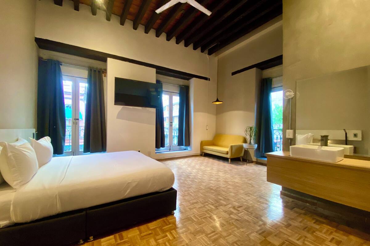 Quarto com cama, sofá, mesa pequena, espelho e banheiro com pia.