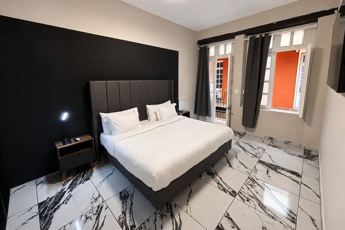 Quarto moderno com uma cama grande, uma mesa de cabeceira, uma luminária no teto e vista para o exterior através de uma janela com cortinas.