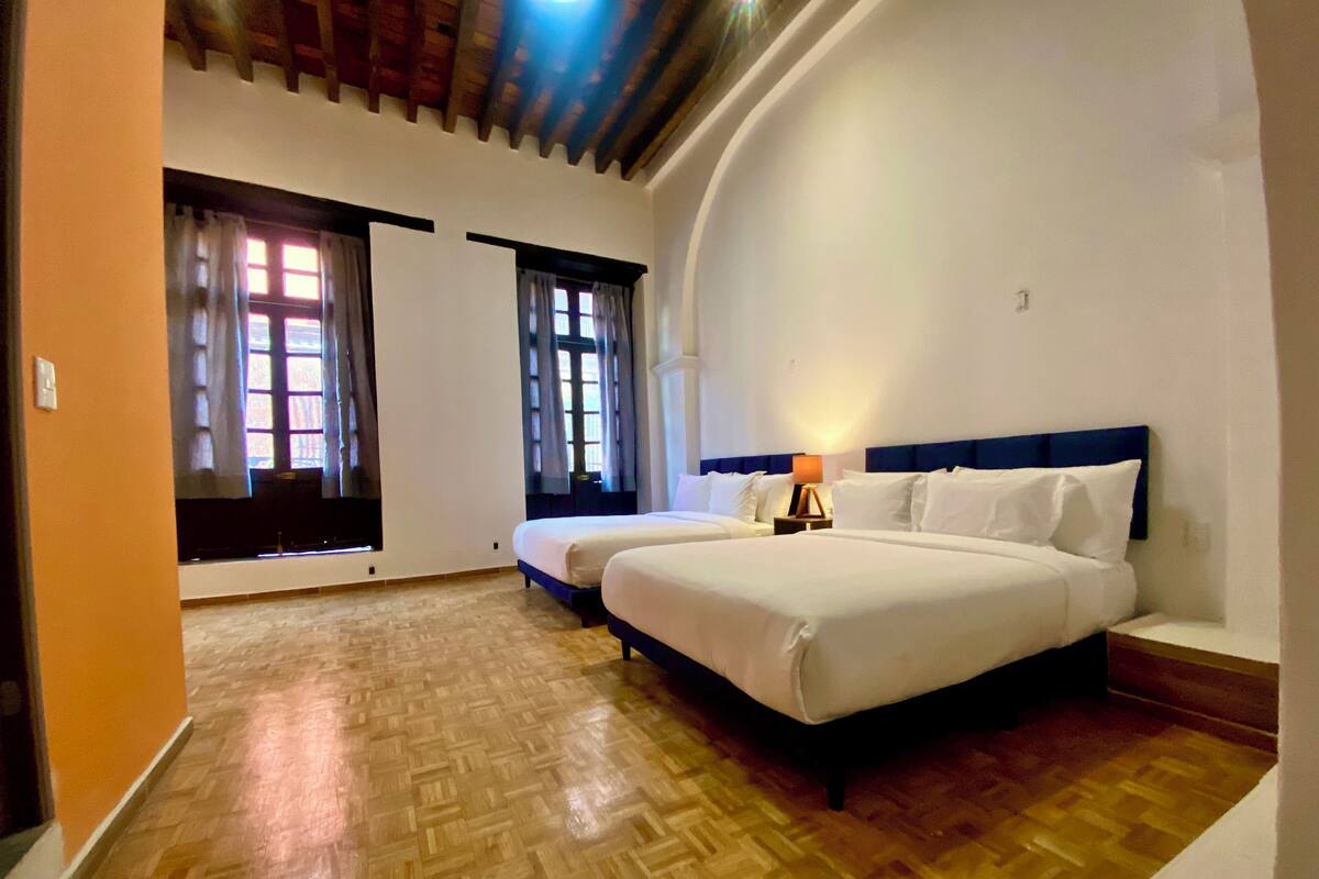 Quarto de hotel com duas camas, piso de madeira, janelas amplas com cortinas e teto com vigas aparentes.