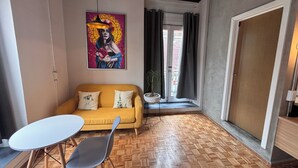 Quarto com sofá amarelo, mesa branca e uma cadeira. Há uma pintura na parede e uma planta perto da janela.