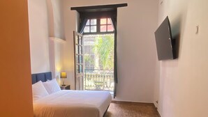 Quarto de hotel com uma cama, mesa de cabeceira com abajur, televisão na parede e vista para uma varanda com plantas.