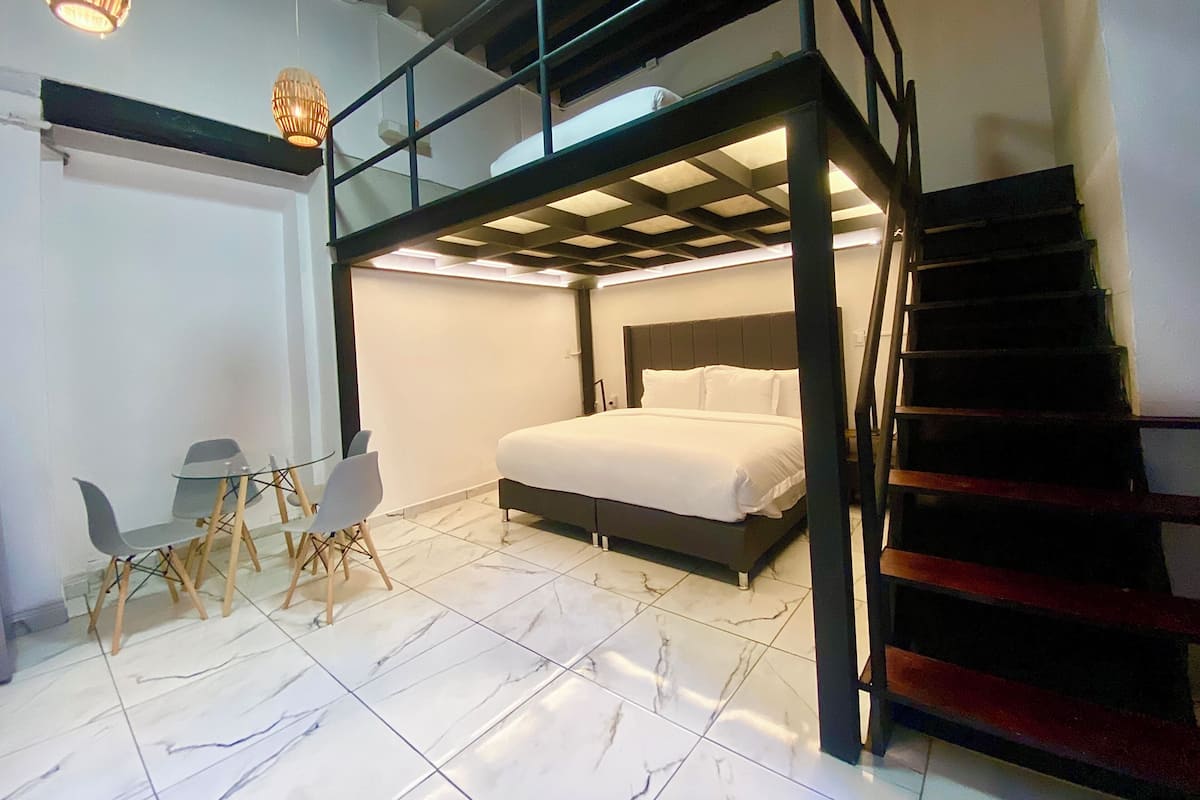 Quarto com cama, escrivaninha com cadeiras, escada e um mezanino com luminárias.