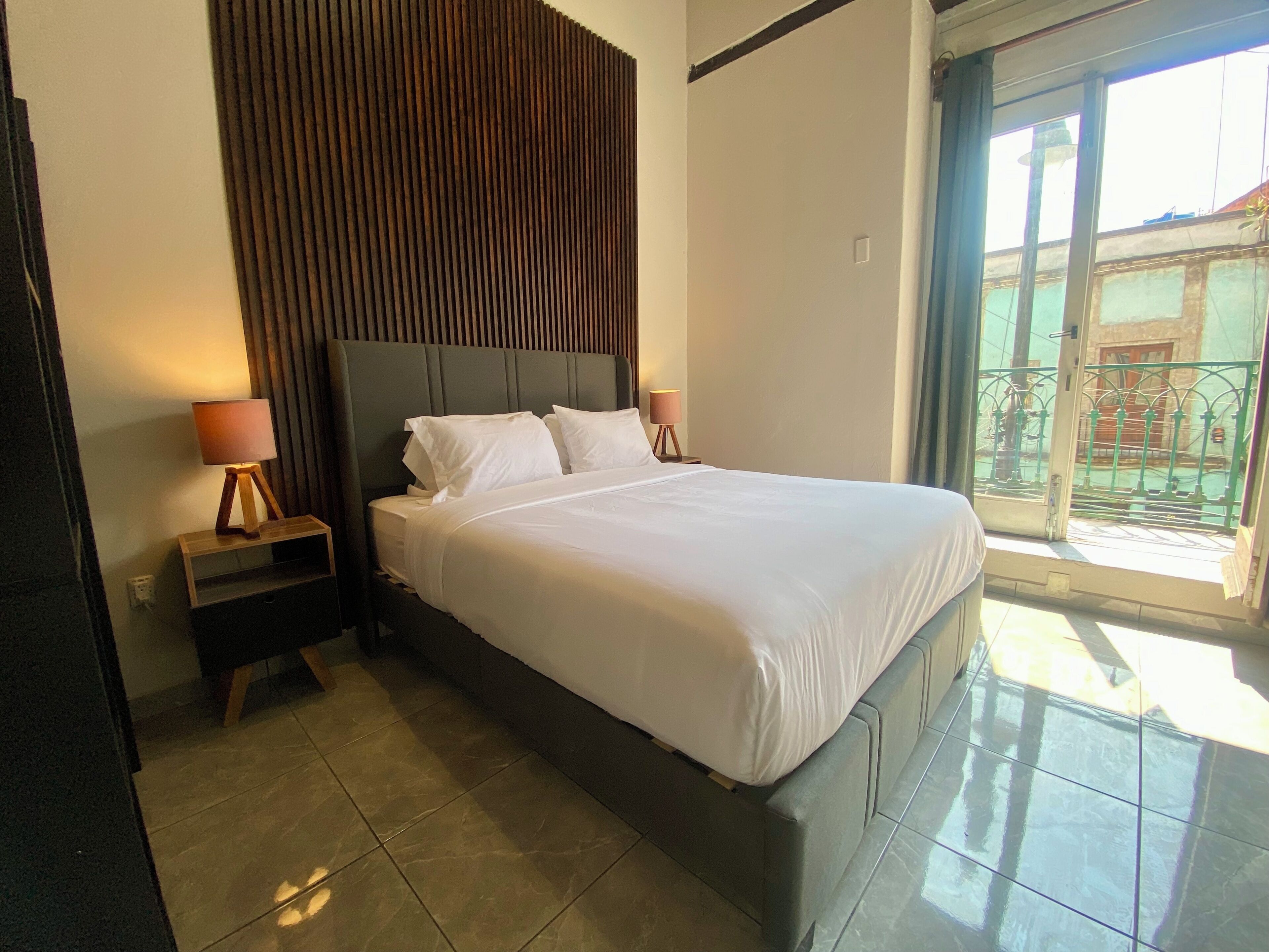 Quarto de hotel com uma cama grande, duas luminárias de cabeceira, um painel de cabeceira em madeira e vista para uma varanda.