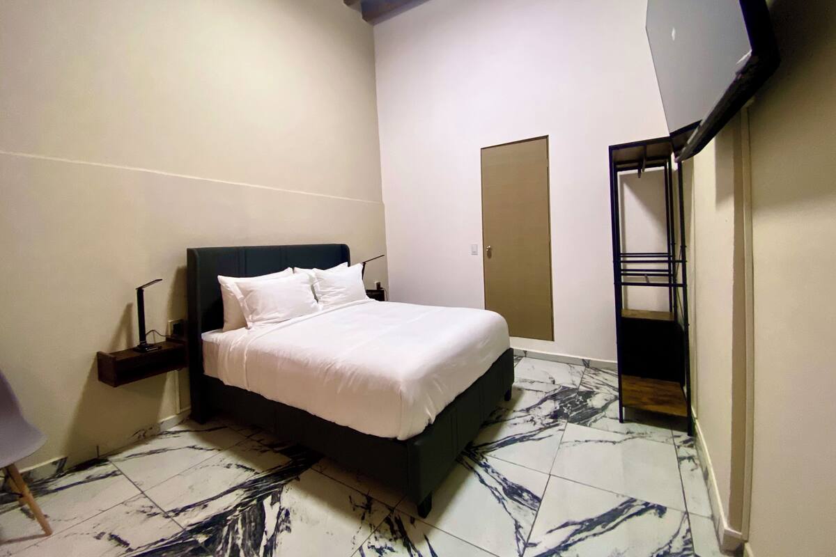 Quarto com cama, mesa de cabeceira, porta e escada.