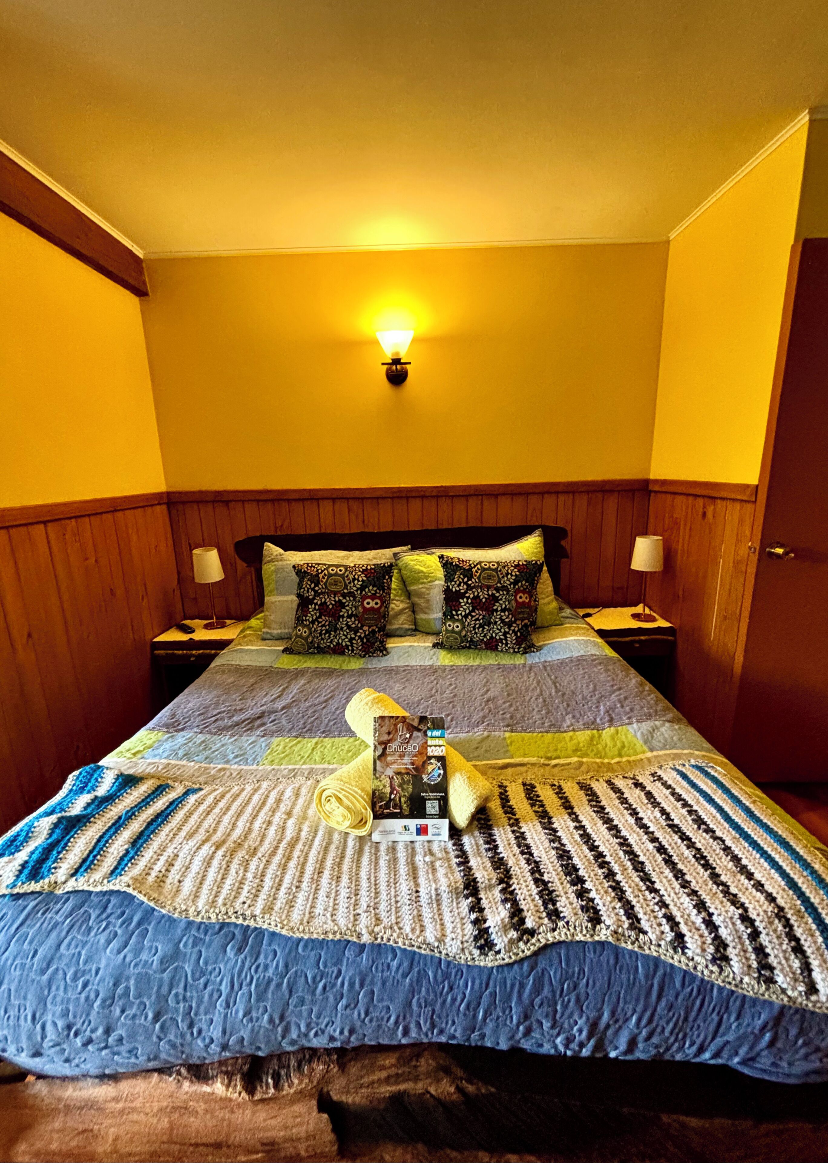 Uma cama com colcha, almofadas, mesa de cabeceira, luminária na parede e uma porta.