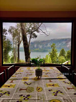 Mesa de jantar com toalha estampada com azeitonas, em frente a uma janela com vista para uma paisagem florestal e um lago.