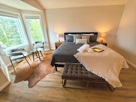 Quarto com uma cama grande, duas luminárias de cabeceira, uma cadeira, uma mesinha e um tapete.