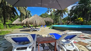 Uma área na beira da piscina com espreguiçadeiras, uma mesa e guarda-sóis de palha.