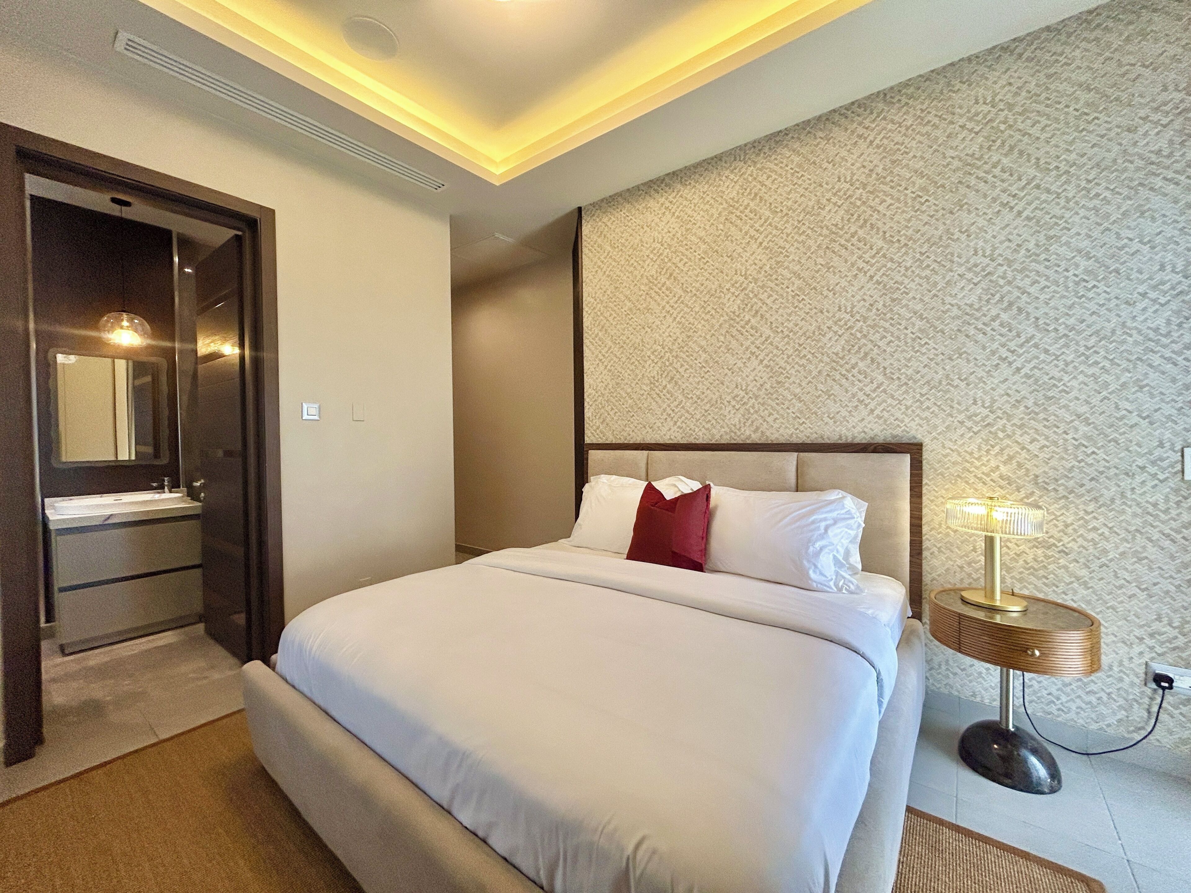 Quarto de hotel com cama, mesa de cabeceira com luminária, espelho e banheiro com pia.