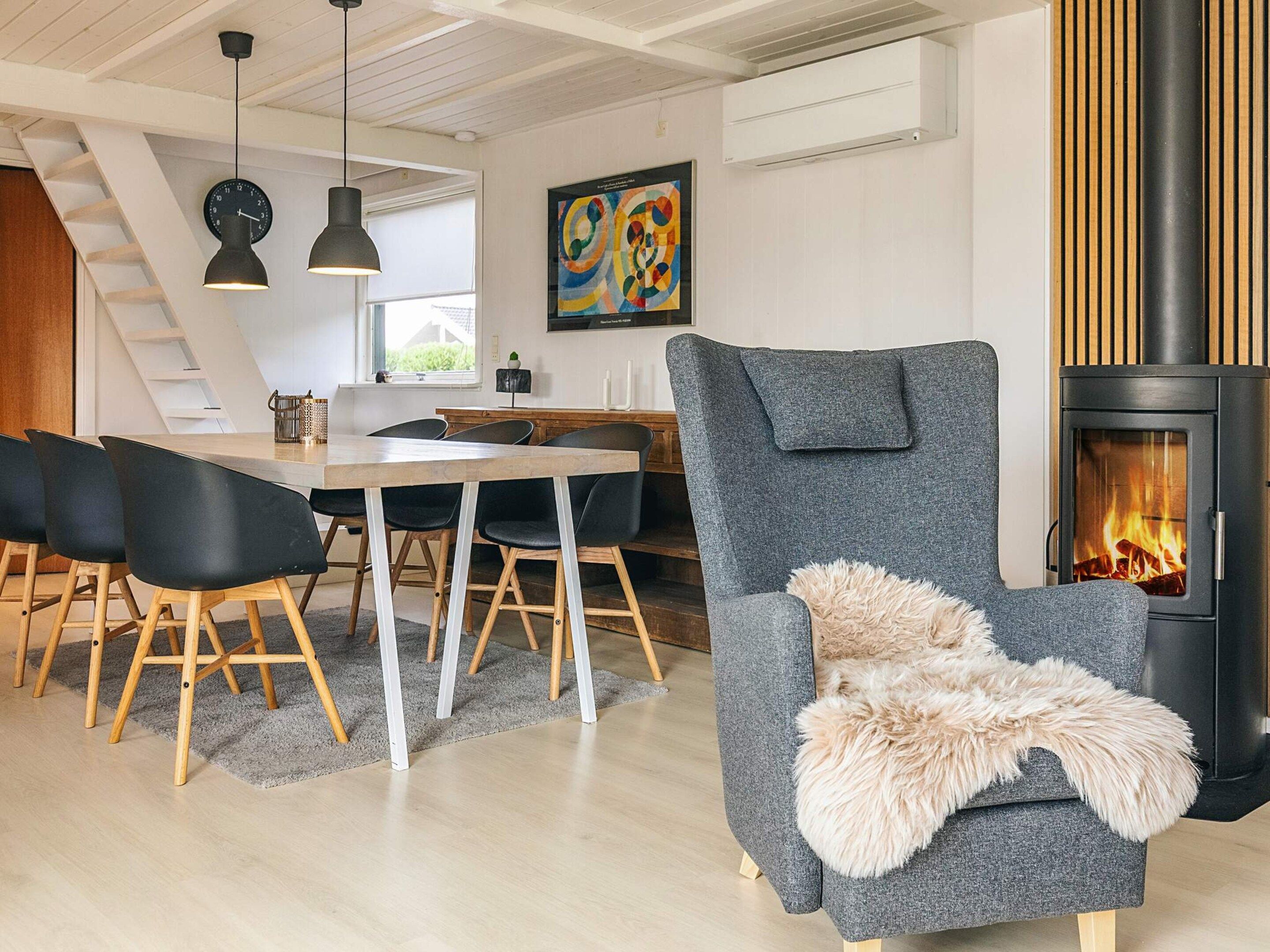 Uma sala de estar moderna com uma poltrona cinza, lareira, área de jantar com mesa e cadeiras de madeira, e escada.