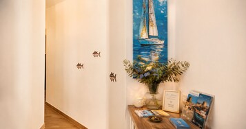 Um corredor com piso de madeira, um quadro de um veleiro, um vaso com flores e uma placa de boas-vindas.