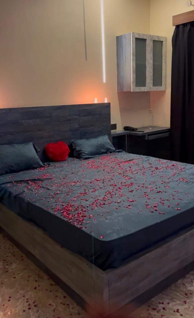 Uma cama com pétalas de rosas espalhadas, uma mesinha de cabeceira com abajur e um armário sobre a cama.