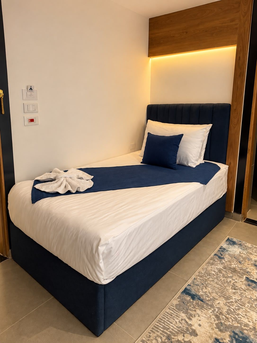 Cama bem arrumada com cabeceira escura, roupa de cama branca e almofada azul decorativa.