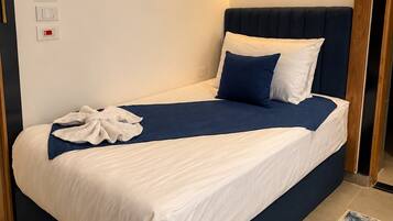 Cama bem arrumada com cabeceira escura, roupa de cama branca e almofada azul decorativa.