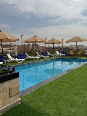 Piscina com espreguiçadeiras e guarda-sóis, cercada por um gramado e paisagem urbana ao fundo.