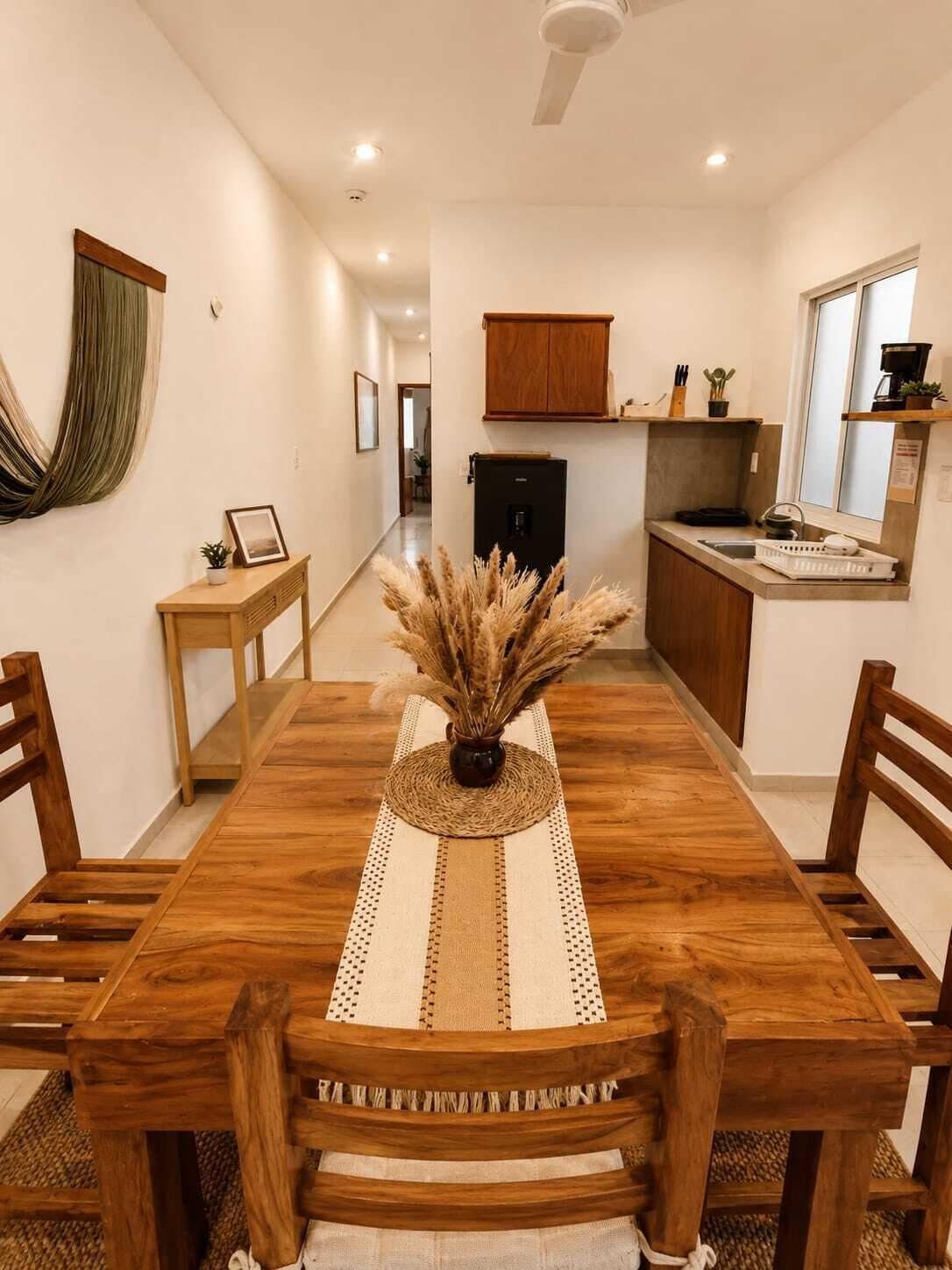 Uma área de refeições com uma mesa de madeira, cadeiras, um vaso com plantas secas e uma cozinha ao fundo.