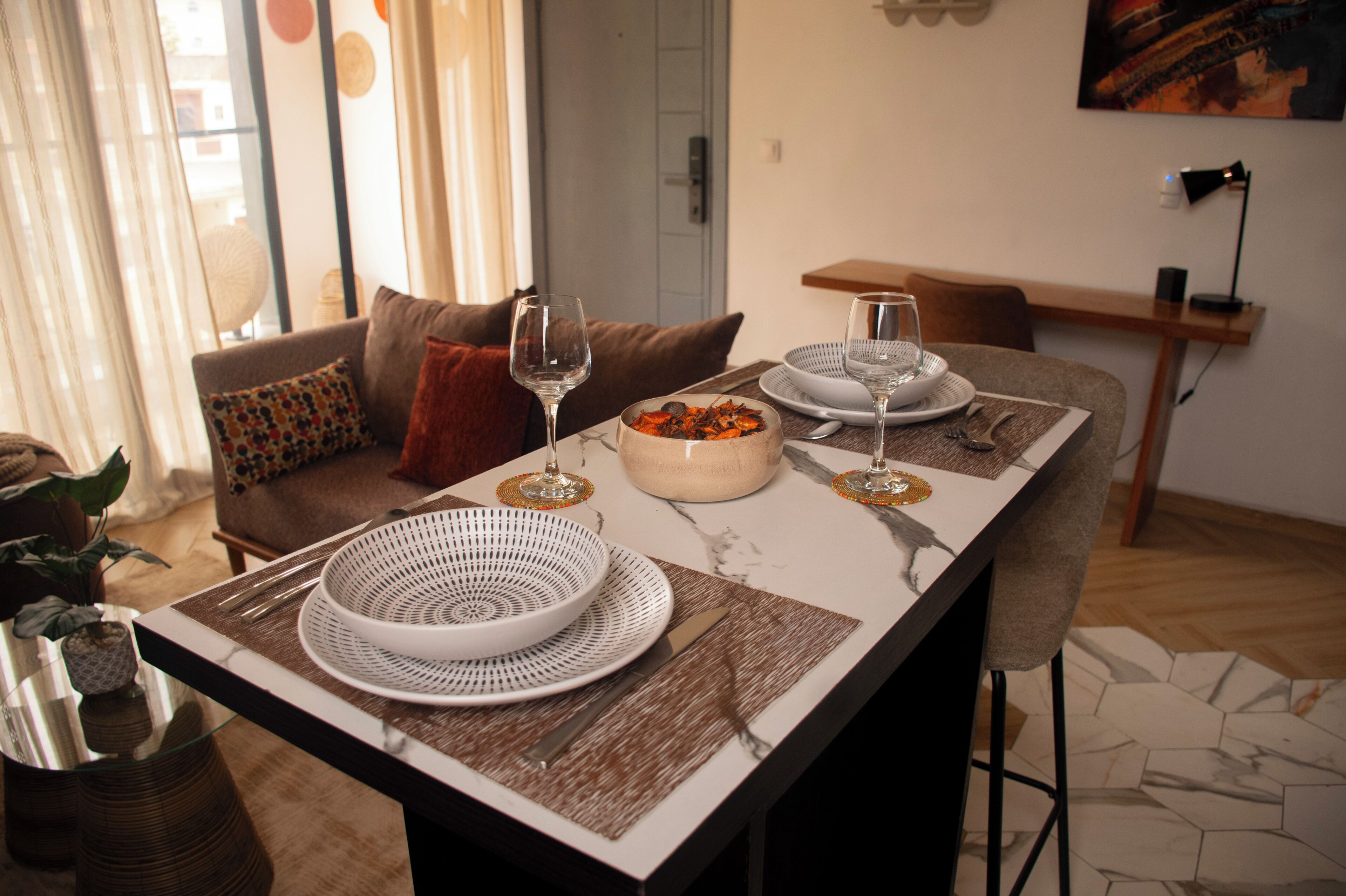 Mesa posta com pratos, tigelas e copos de vinho, um sofá com almofada estampada e um vaso com planta.